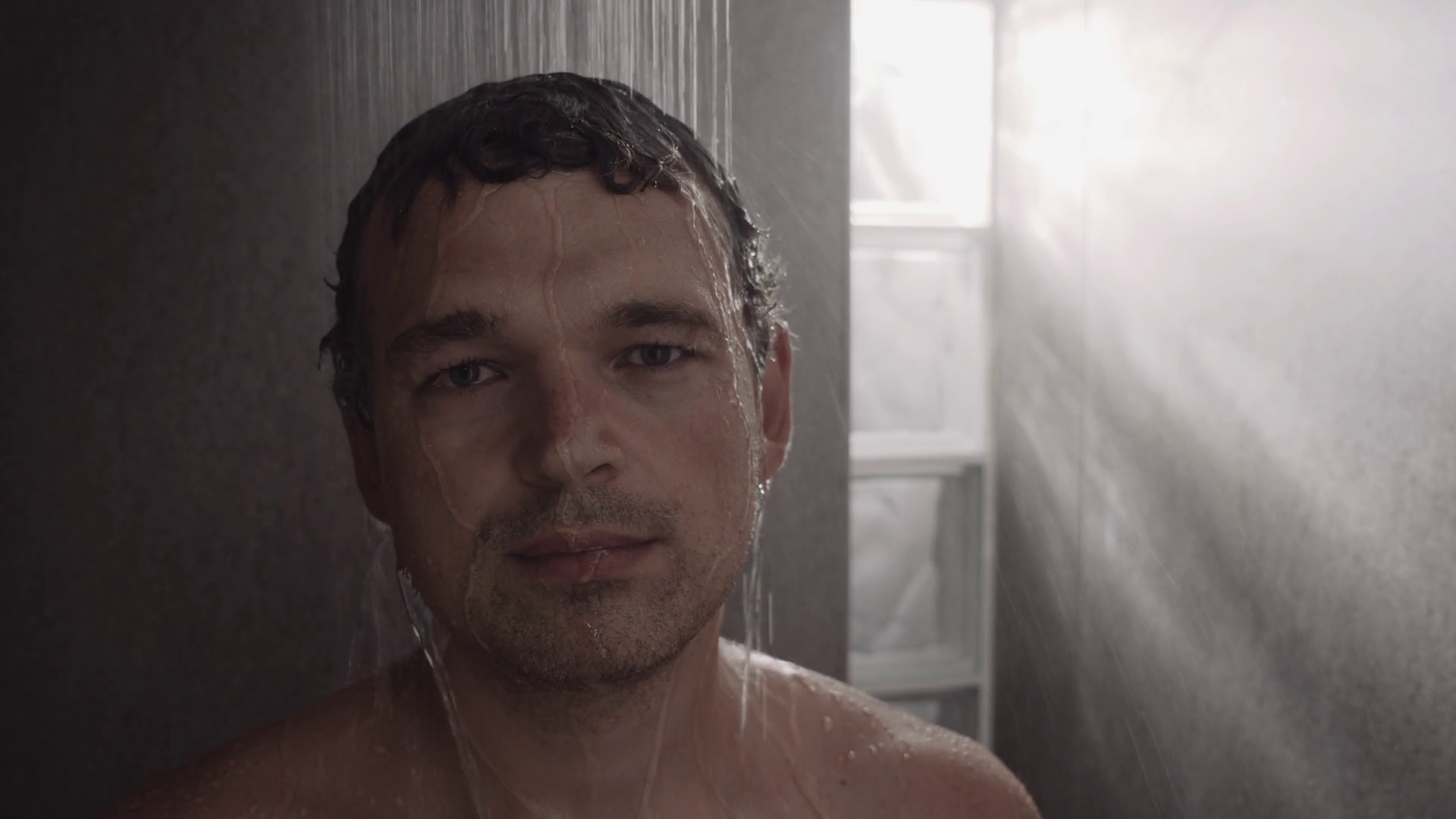 A portrait of a young sad man, standing in a shower under water spray ...