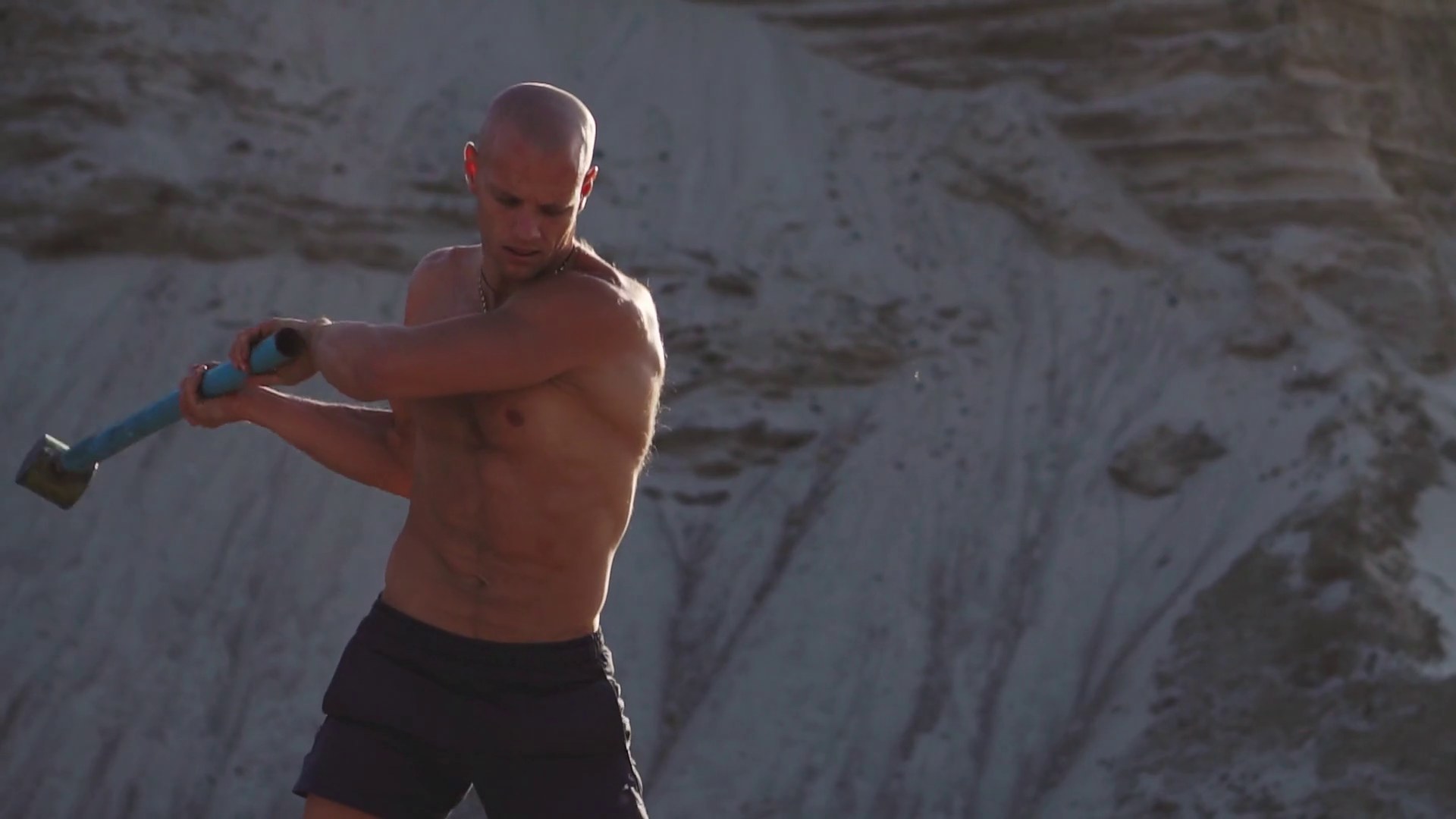 Bald man strongman hits a hammer on a huge wheel in the sandy mountains ...