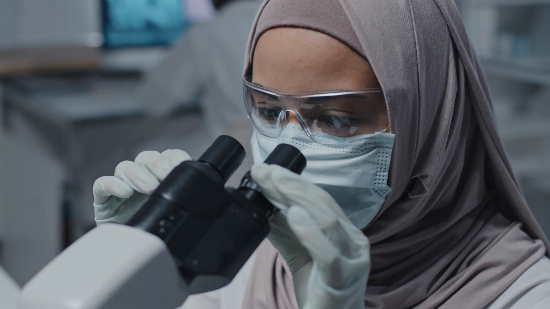 Close-up slow motion of young Muslim female lab technician in gloves, mask and goggles sitting ...
