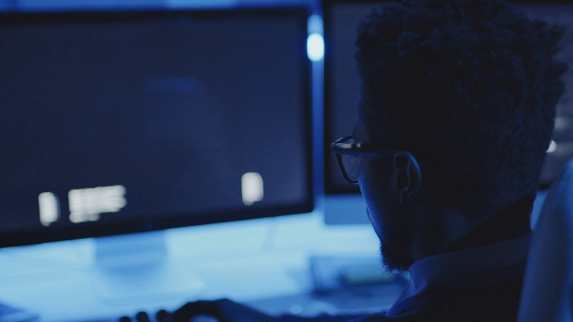 Over-shoulder of young Black male programmer sitting at desk in dark ...